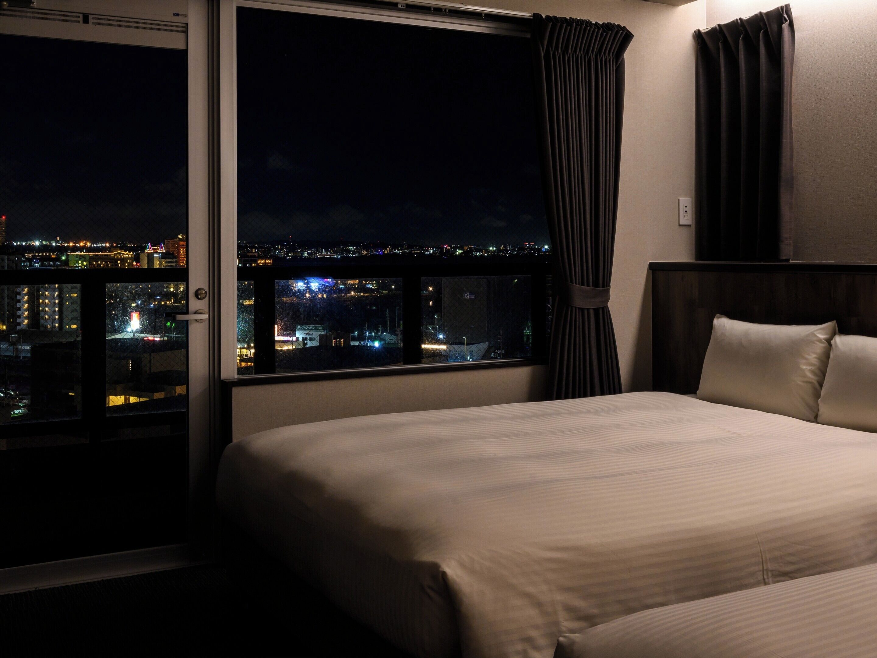 Quarto de hotel com cama, janela com cortinas e vista para a paisagem urbana noturna.