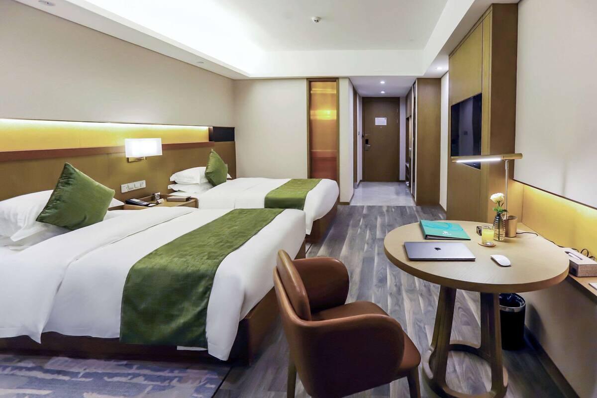 Quarto de hotel com duas camas, uma escrivaninha, uma cadeira, um laptop e uma mesinha de cabeceira com abajur.
