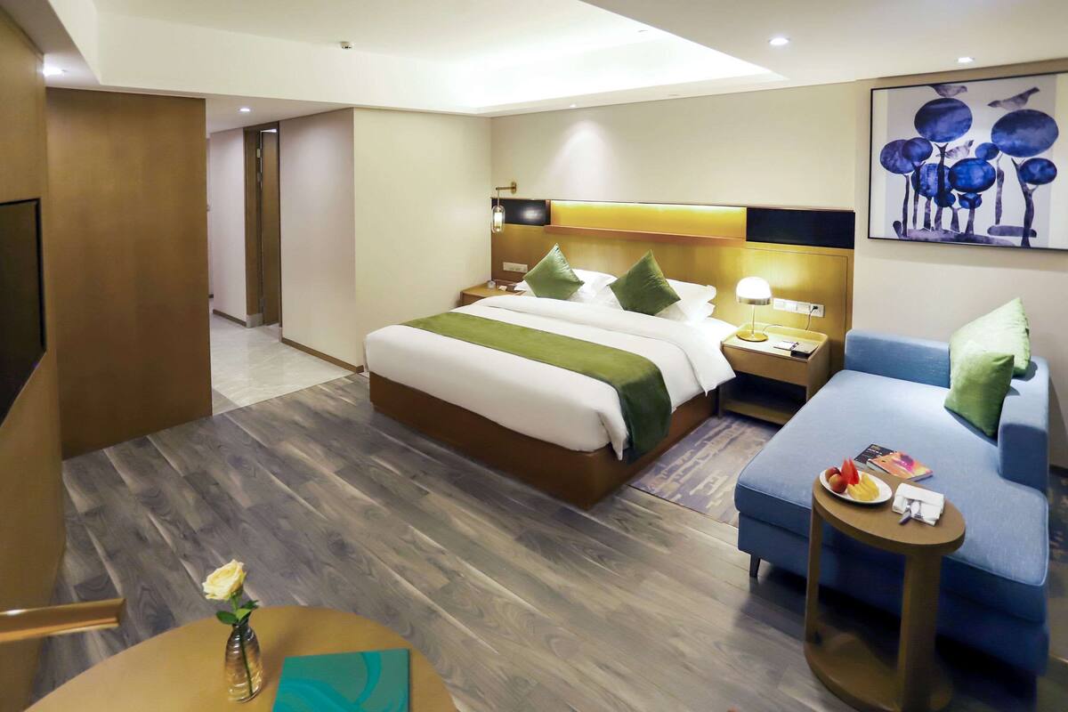 Quarto de hotel com cama, mesas de cabeceira, escrivaninha, cadeira e uma pequena mesa com um vaso de flores.