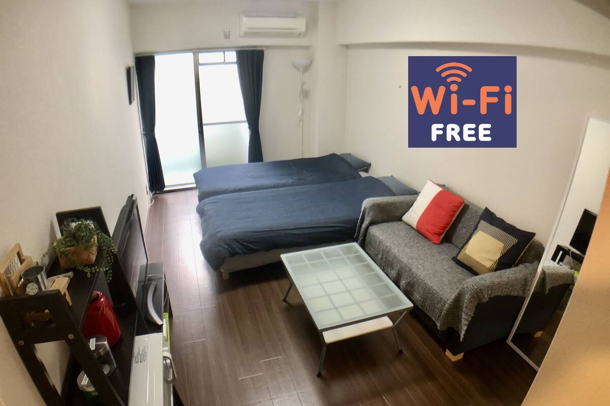 Quarto de hotel com cama, sofá, mesinha, televisão, planta e um aviso indicando Wi-Fi gratuito.