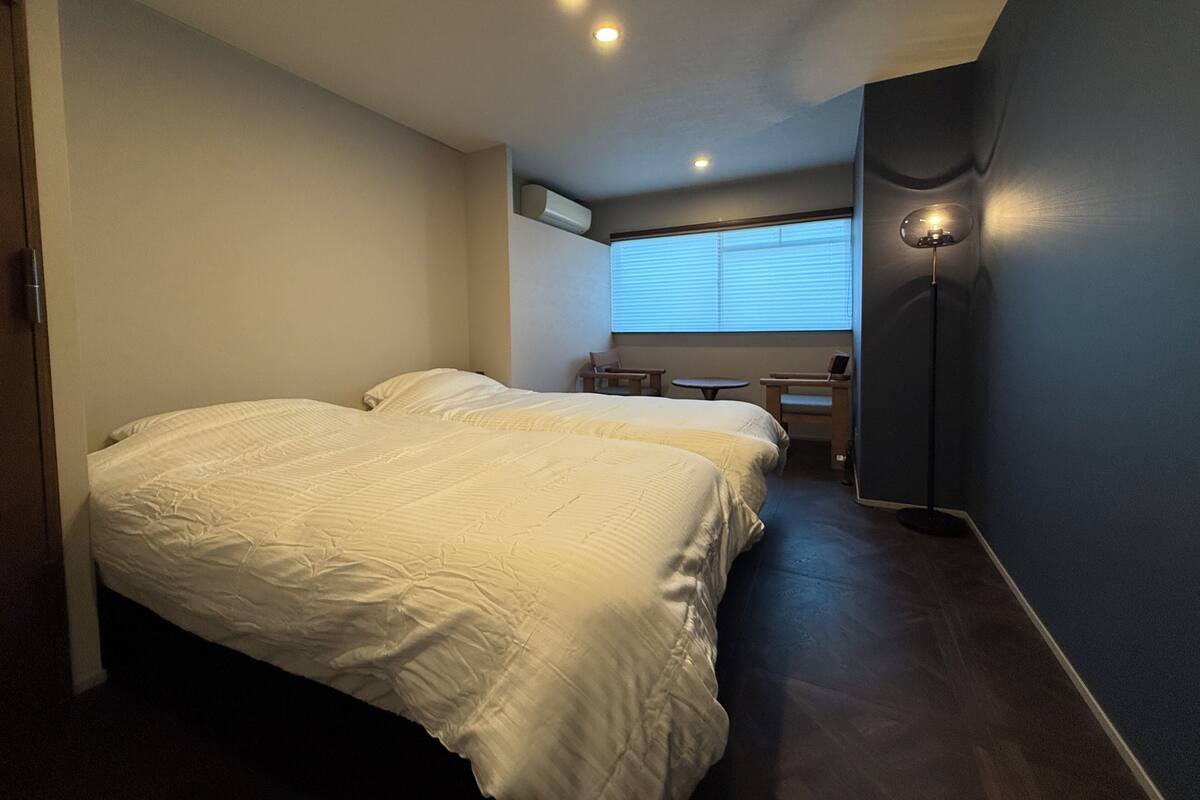 Quarto de hotel com duas camas, uma escrivaninha, uma cadeira, um abajur e uma janela com persianas.