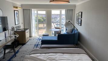 Quarto de hotel com cama, escrivaninha, cadeira, sofá, mesa de jantar e vista da cidade através de janelas amplas.