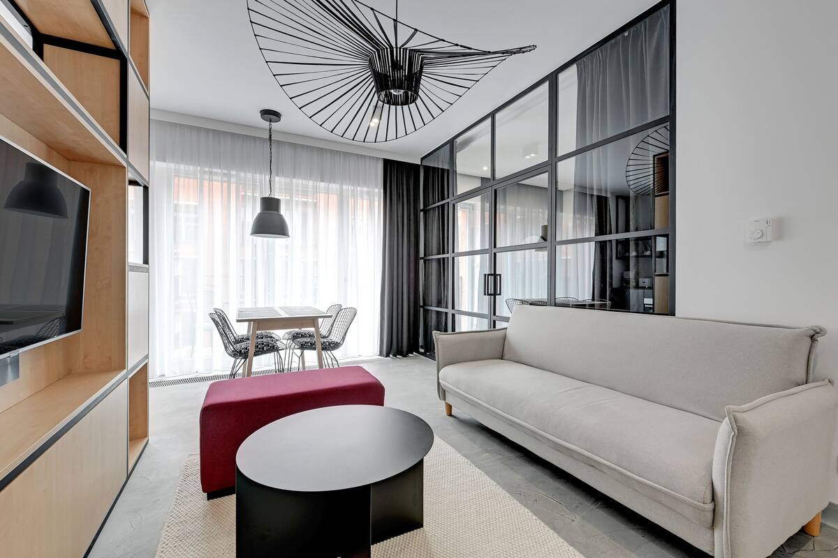 Uma sala de estar moderna com um sofá, um banquinho vermelho, uma mesa de centro redonda e uma TV em uma estante de madeira.