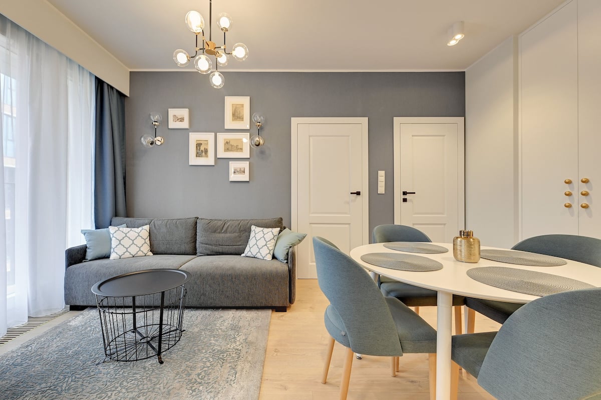 Uma sala de estar moderna com um sofá cinzento, uma mesa de centro redonda e uma mesa de jantar com cadeiras. Há quadros nas paredes e um candelabro sobre a mesa de centro.