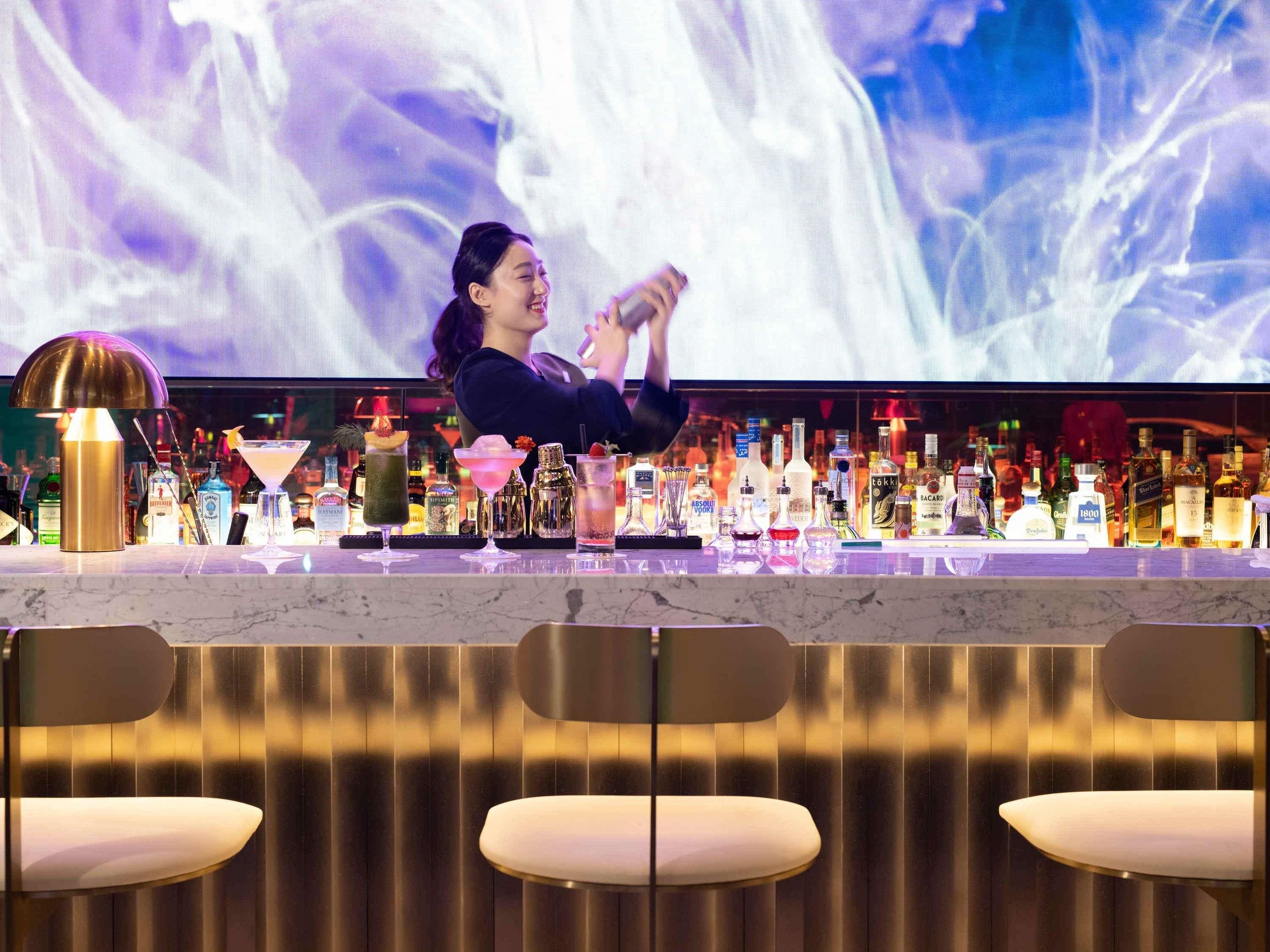 Uma área de bar com um bartender preparando bebidas, diversas garrafas de licor e uma tela grande ao fundo.