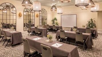 Sala de conferências com mesas preparadas para reuniões, equipada com cadeiras, mesas e plantas decorativas.