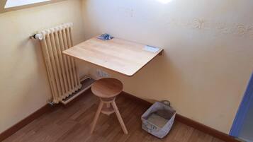 Um quarto pequeno com uma escrivaninha de madeira, um banquinho, um radiador e uma lixeira.