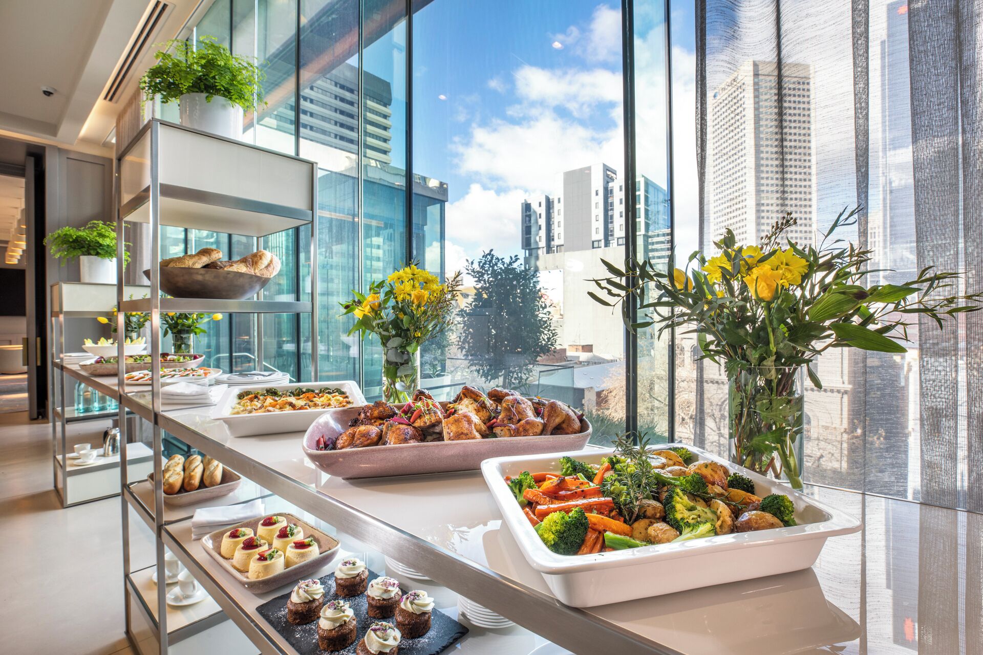 Buffet com diversos pratos, incluindo frango assado, legumes e sobremesas, montado em quarto de hotel moderno com amplas janelas e vista para a cidade.