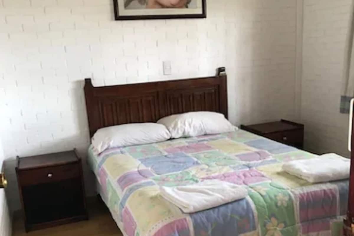 Um quarto com cama de madeira, almofadas brancas, colcha colorida, mesa de cabeceira e um quadro na parede.
