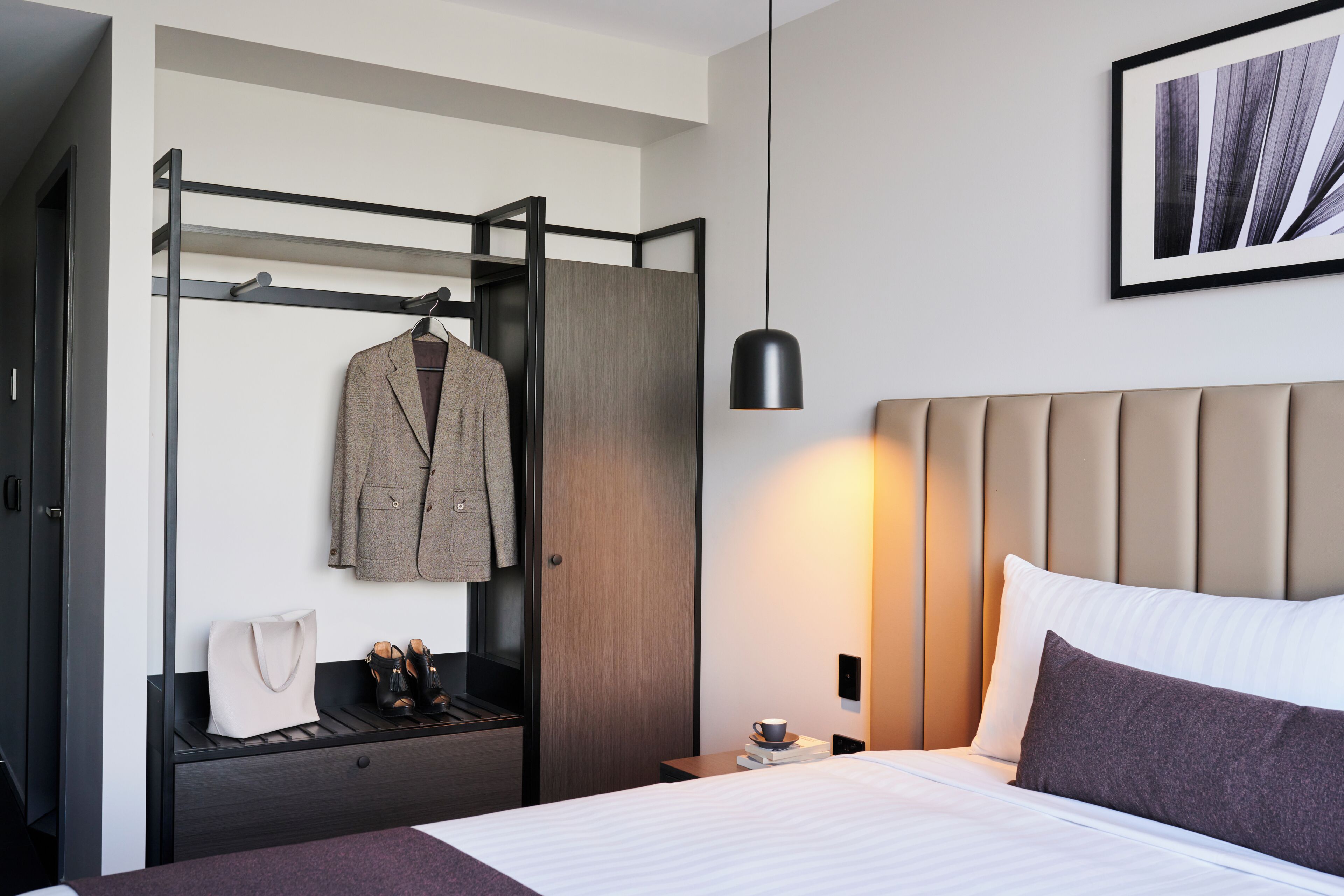 Quarto de hotel com cama, guarda-roupa, cabideiro, mesa de cabeceira com uma chávena e um livro, e uma luz pendente.