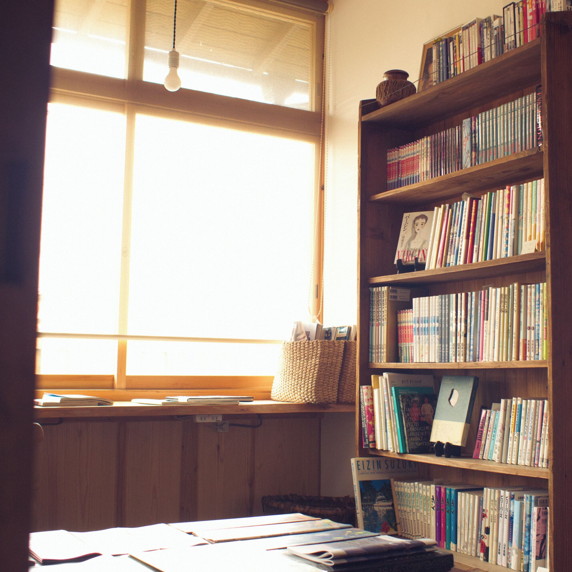 Um quarto com uma grande janela, uma estante de madeira repleta de livros e uma pequena mesa com livros e uma luminária.