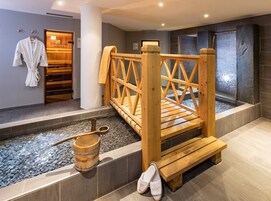 Um banco de sauna de madeira com um balde de madeira e chinelos, outro banco de madeira e a entrada da sauna com um porta-toalhas.