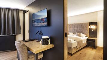 Quarto de hotel moderno com escrivaninha de madeira, cama com cabeceira, televisão fixada na parede e mesa de cabeceira com abajur.
