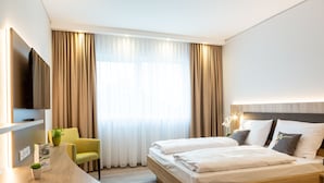 Quarto de hotel com cama de madeira, uma mesa, uma cadeira, televisão e janela com cortinas.