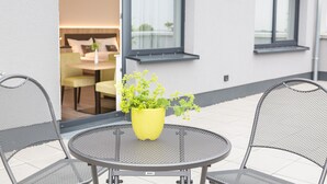 Uma área de pátio com uma mesa redonda de metal e duas cadeiras de malha, uma planta em vaso amarelo e um vislumbre de um espaço de estar interno com um sofá e uma mesa.