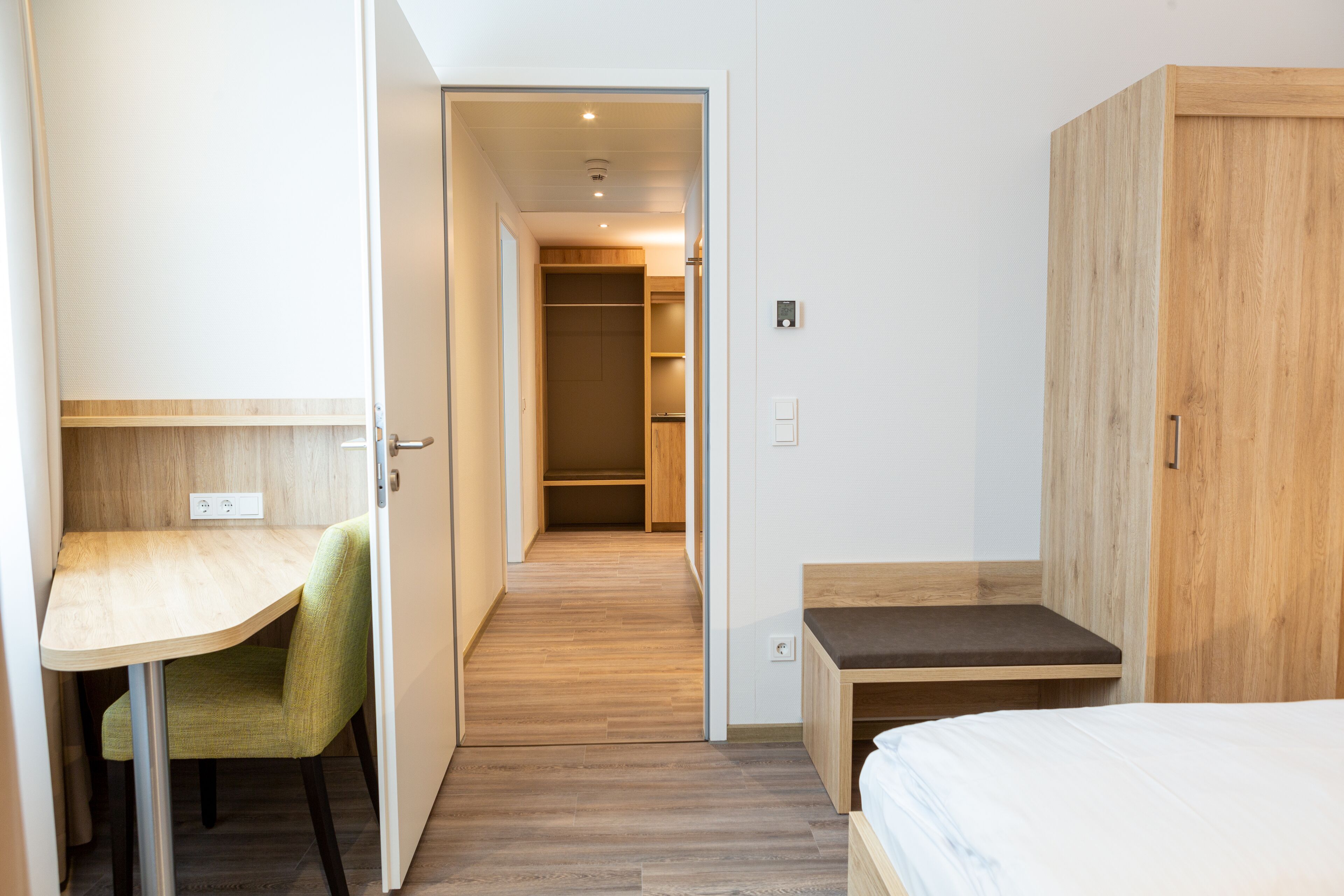 Quarto de hotel compacto com cama, escrivaninha com cadeira, guarda-roupa e um pequeno espaço para sentar.
