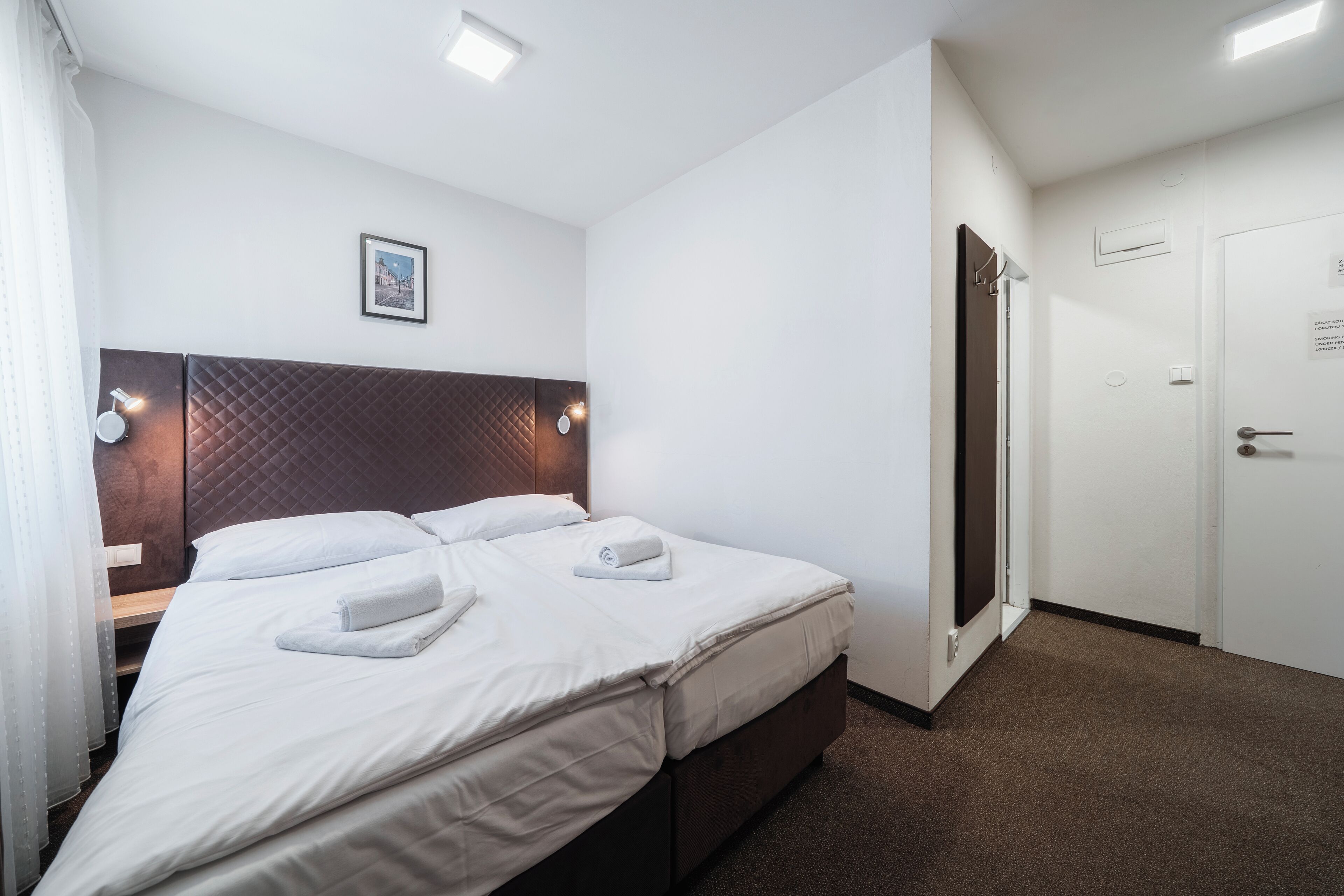 Quarto de hotel com cama, cabeceira, mesa de cabeceira, luminária na parede, quadro emoldurado e porta com placa.