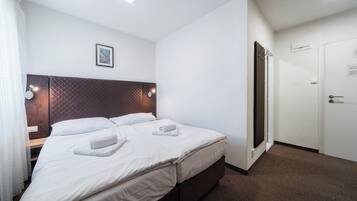 Quarto de hotel com cama, cabeceira, mesa de cabeceira, luminária na parede, quadro emoldurado e porta com placa.