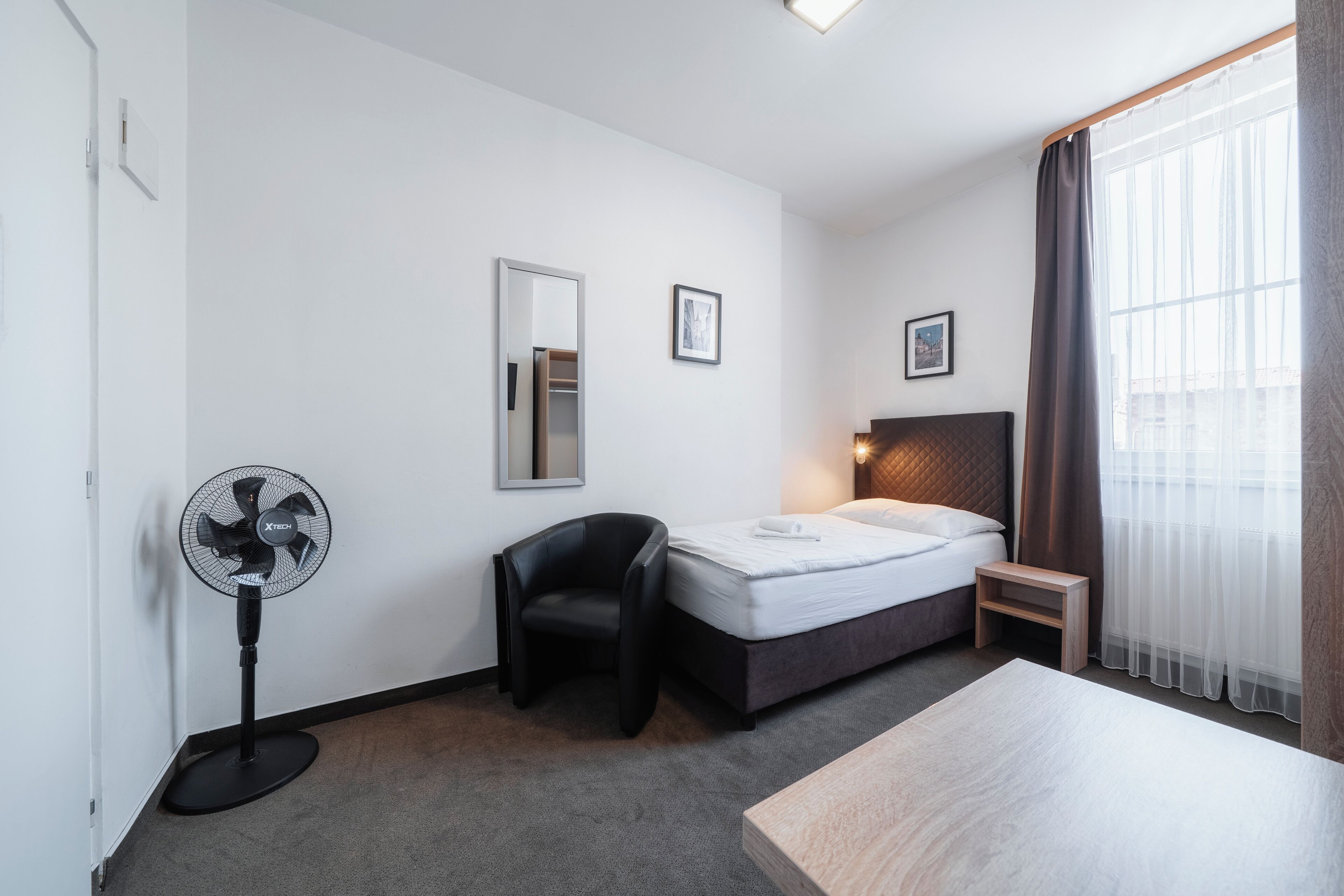 Quarto de hotel com cama, cadeira, mesinha, ventoinha e janela com cortinas.