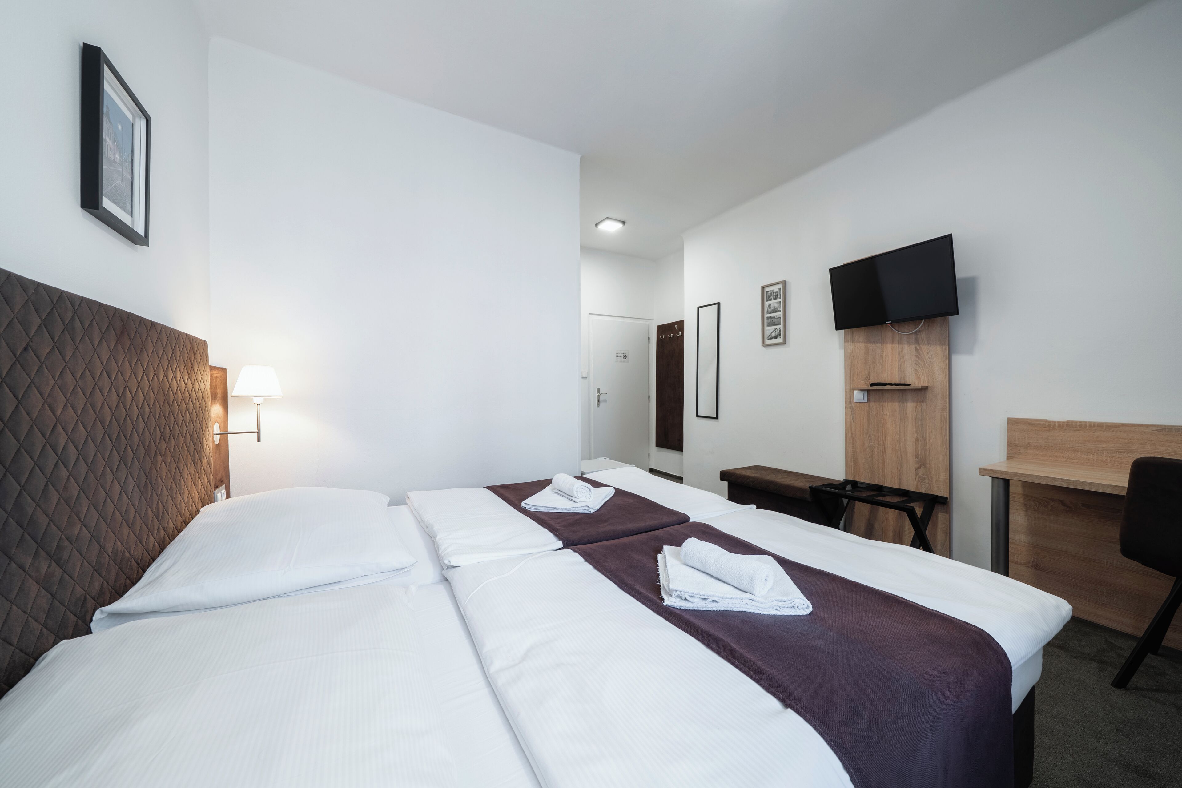 Quarto de hotel com uma cama grande, uma escrivaninha, uma cadeira, uma TV fixada na parede e um quadro emoldurado.