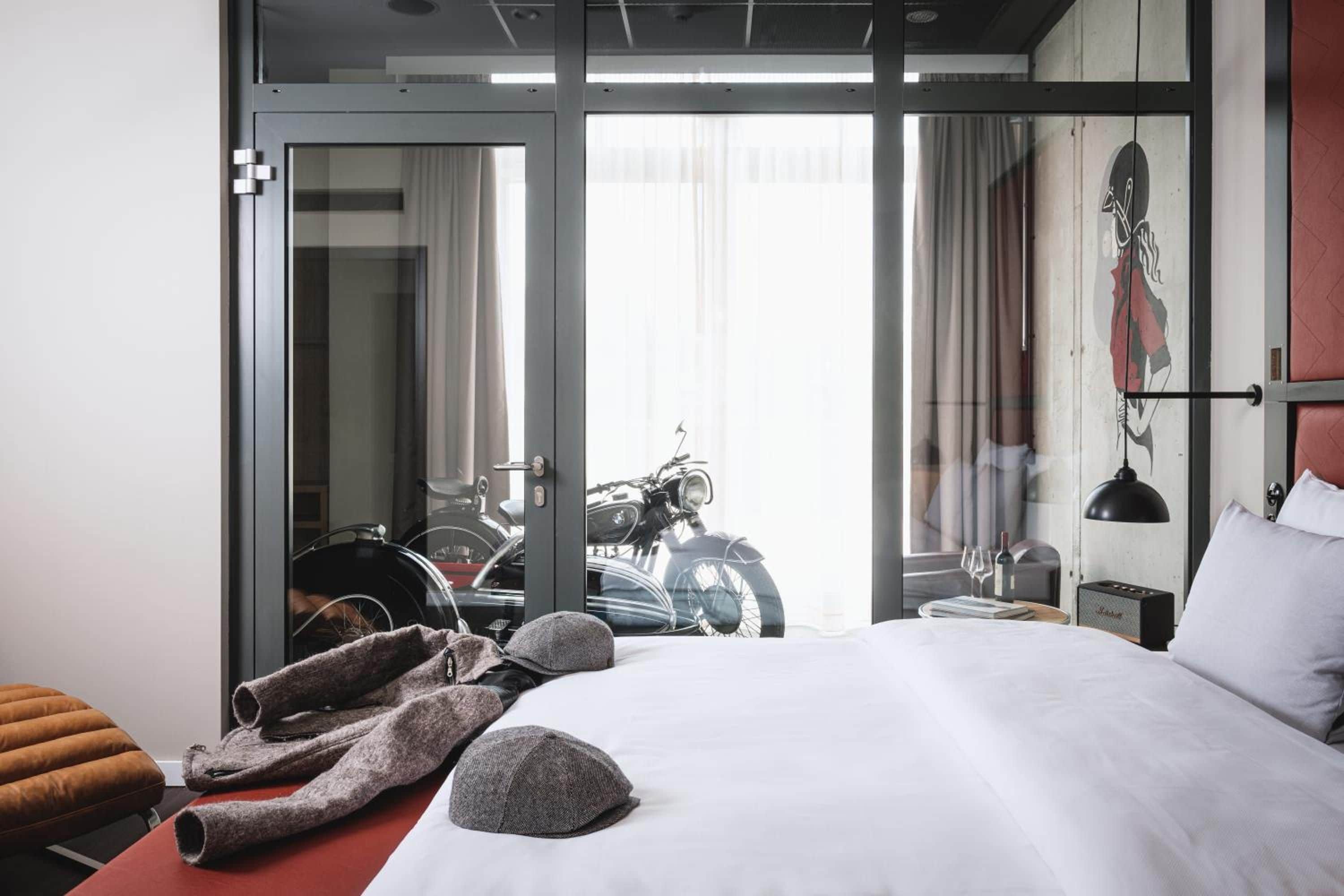 Quarto de hotel com cama, um banquinho vermelho, uma motocicleta e uma janela grande.