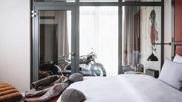 Quarto de hotel com cama, um banquinho vermelho, uma motocicleta e uma janela grande.