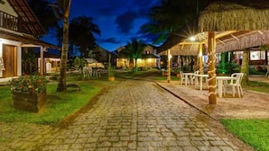 Cena noturna de um resort com um caminho de pedras, construções com telhados de palha e áreas de estar ao ar livre.