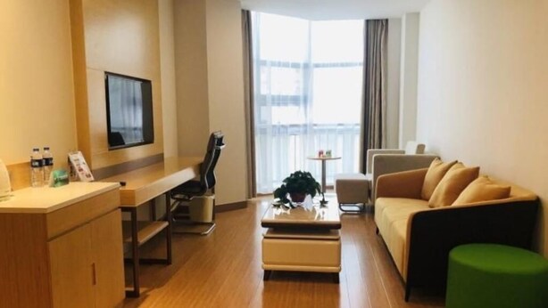 Quarto de hotel moderno com piso de madeira, um sofá, um banquinho verde, uma mesa de centro, uma TV e uma pequena mesa com uma planta.