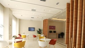 Um hall de hotel moderno com mesas e cadeiras redondas, um balcão de recepção e uma tela de televisão.