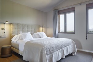 Cama bem arrumada com colcha branca texturizada, duas luminárias de cabeceira, mesa de cabeceira redonda e janela com cortinas.