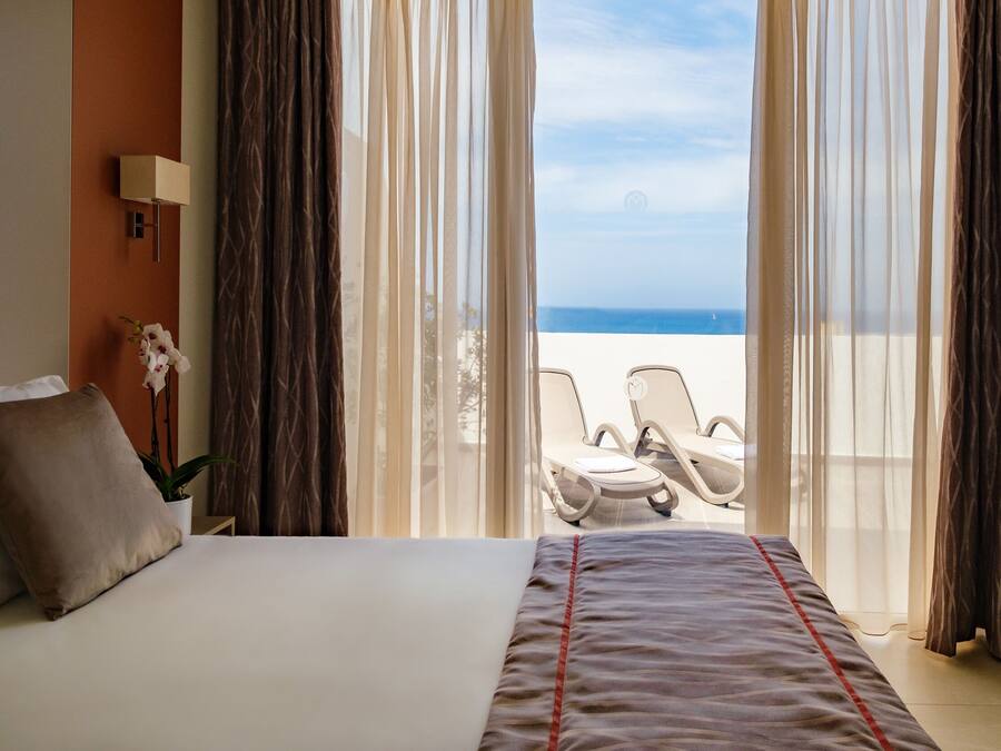 Quarto de hotel com cama, planta em vaso, escrivaninha e vista para a praia.
