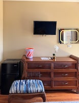 Quarto com cômoda de madeira, televisão fixada na parede, mesa pequena com um vaso e um banco.