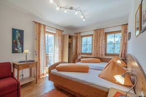 Uma cama de madeira com duas almofadas, uma mesa de cabeceira com abajur, uma cadeira vermelha e uma janela com cortinas.