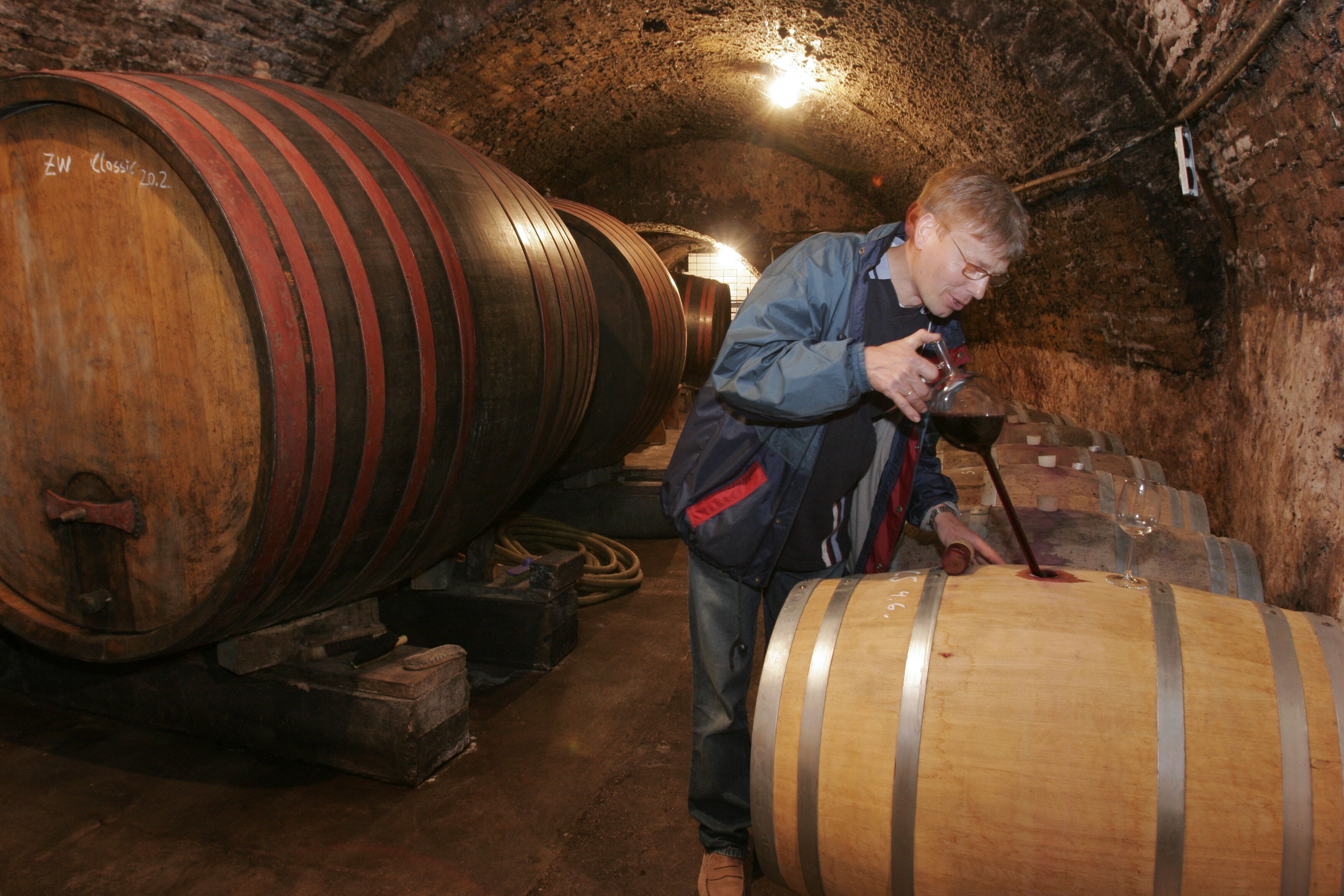 Uma pessoa servindo vinho de um barril em adega com grandes barris de madeira e parede de pedra.