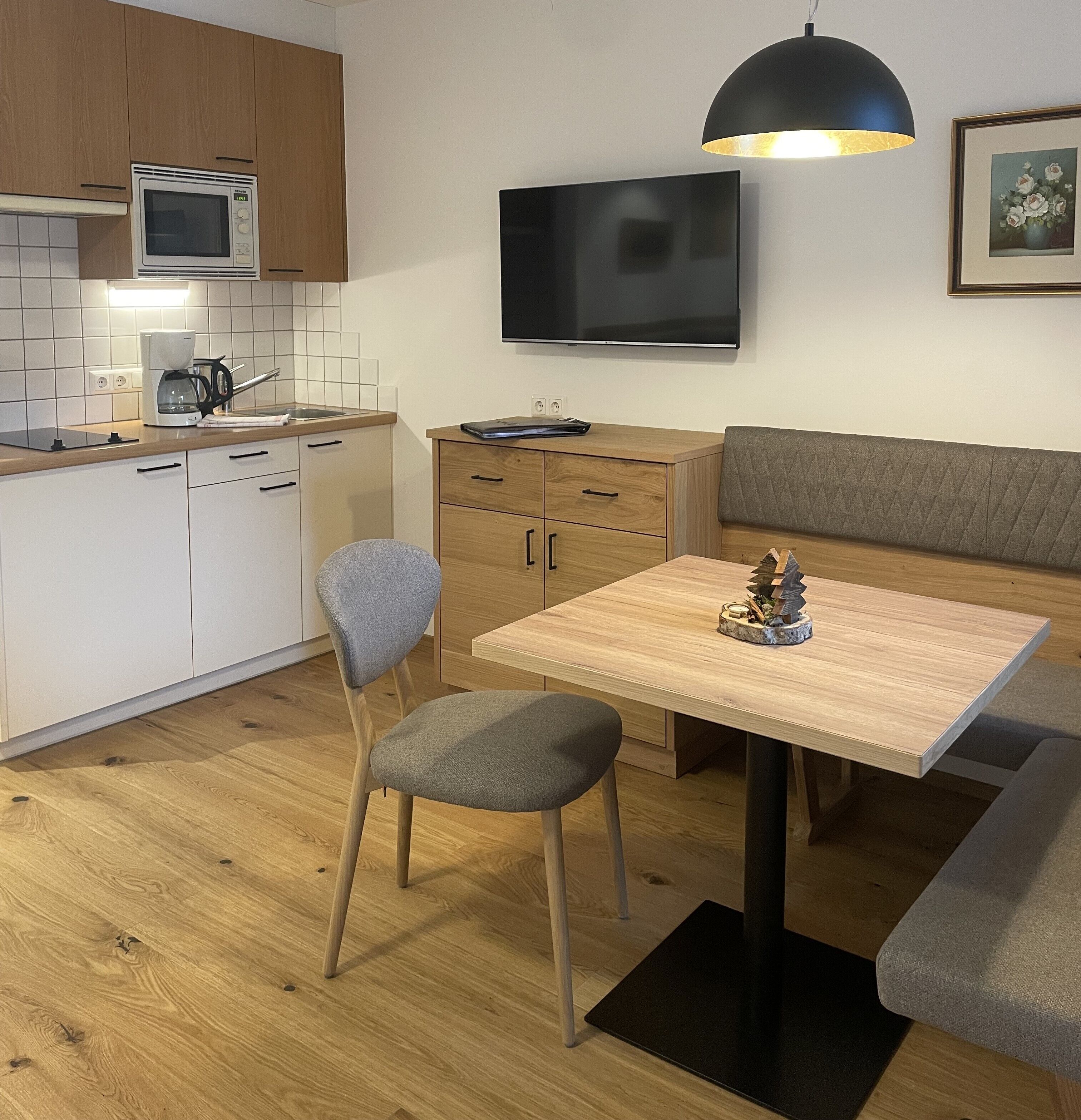 Uma cozinha compacta com micro-ondas, cafeteira e TV de tela plana. Há uma mesa de jantar com um centro de mesa decorativo e um banco.