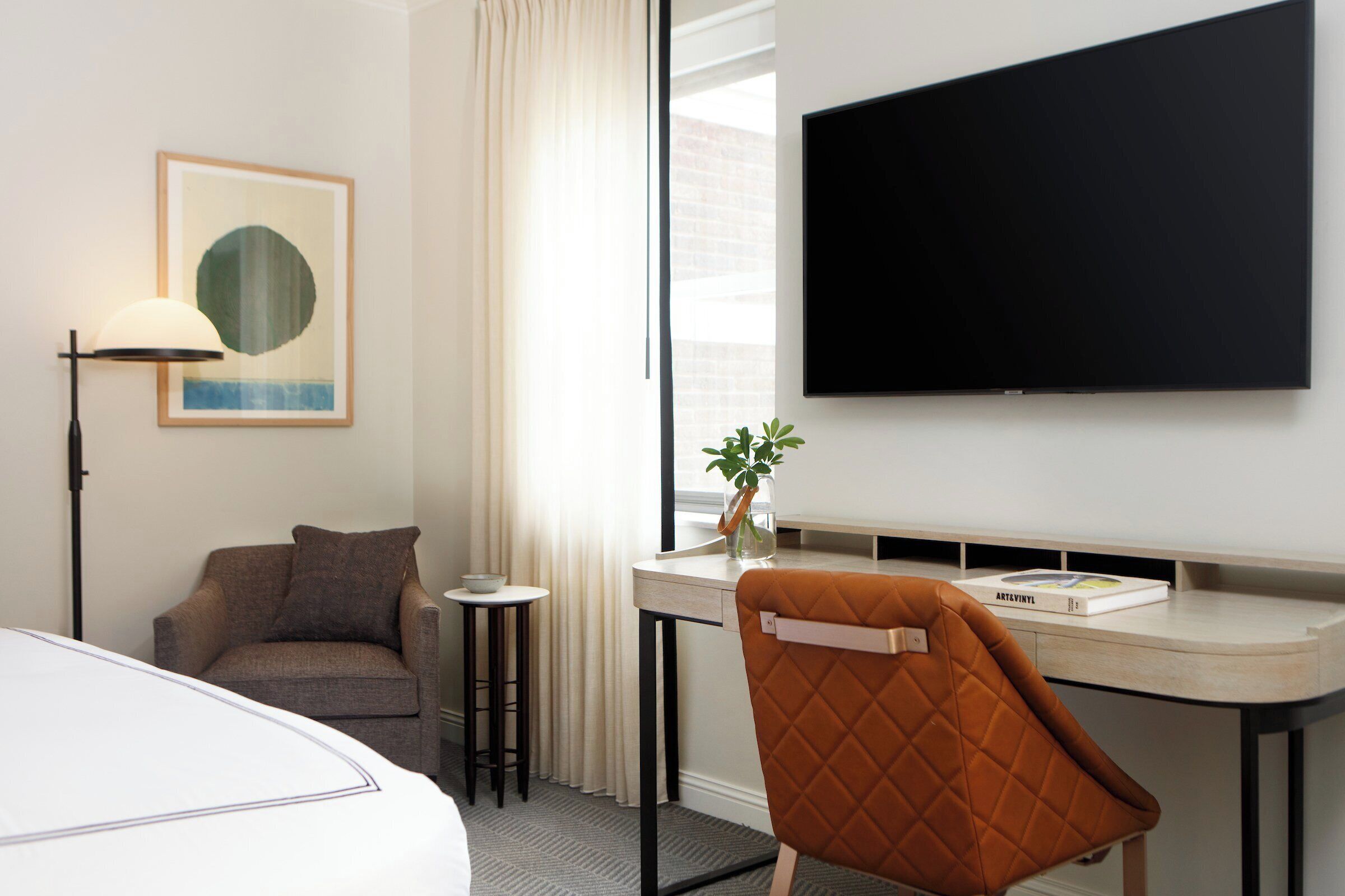 Quarto de hotel com cama, escrivaninha com cadeira, televisão, luminária, pintura e janela com cortinas.