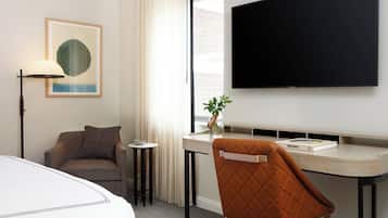Quarto de hotel com cama, escrivaninha com cadeira, televisão, luminária, pintura e janela com cortinas.
