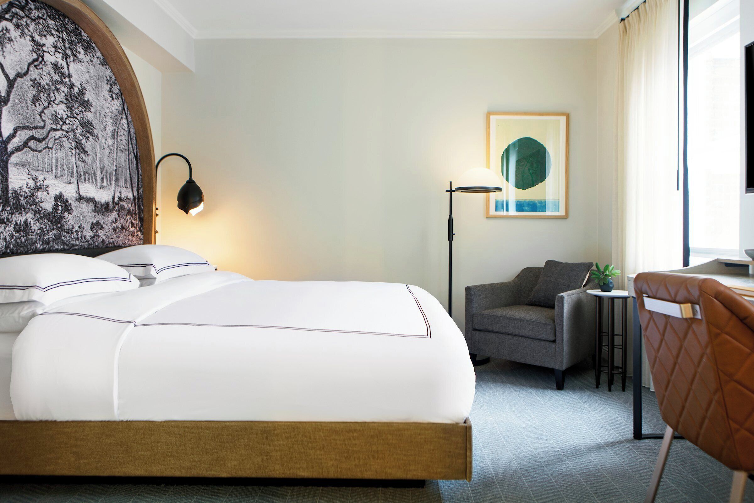 Quarto de hotel com cama, escrivaninha, cadeira, luminária de chão e um quadro na parede.