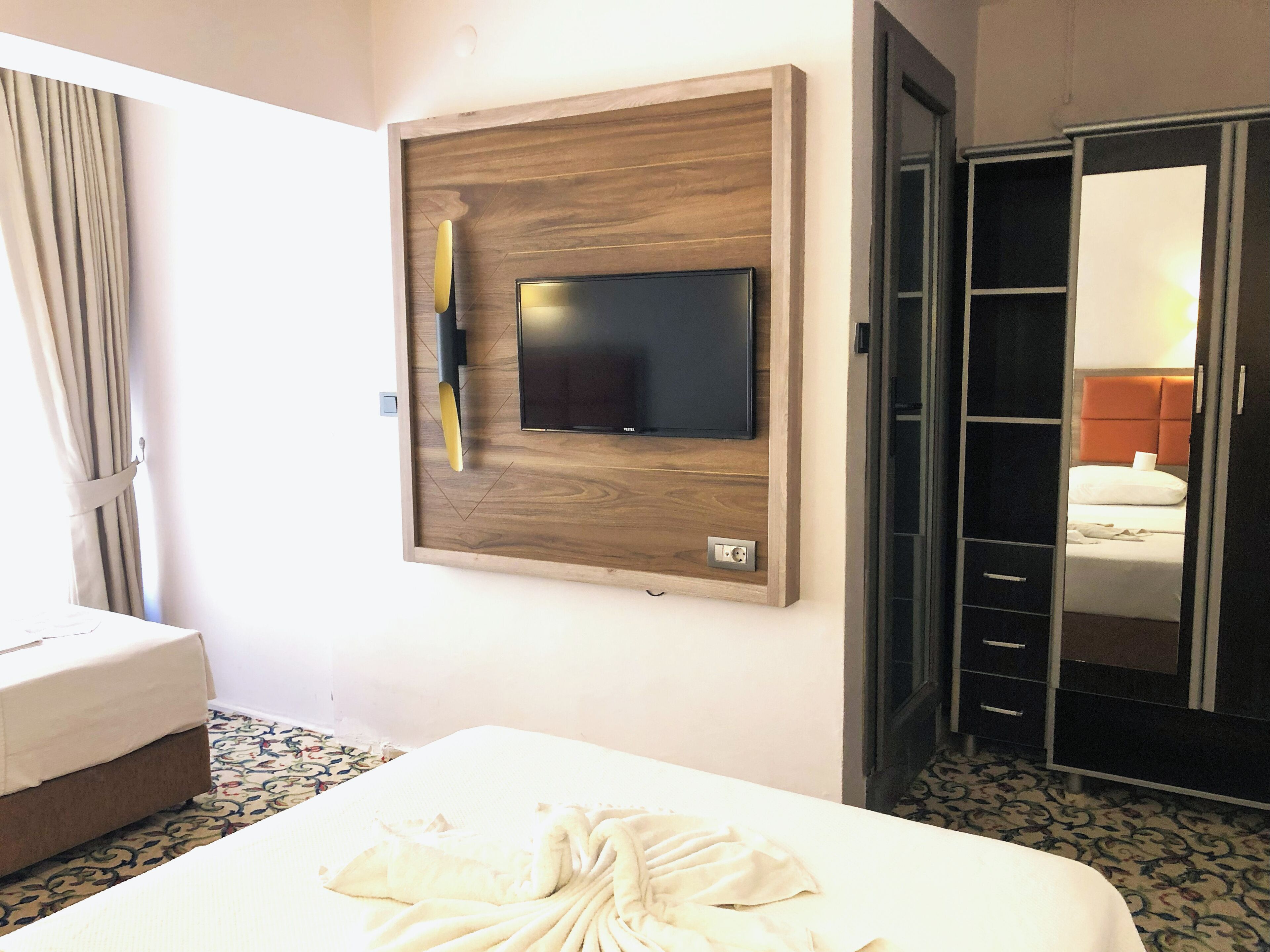 Quarto de hotel com cama, televisão presa a um painel de madeira, guarda-roupa e banheiro com cortina.