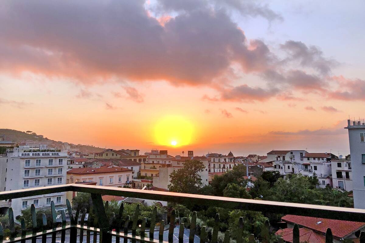Vista do pôr do sol a partir de uma varanda com vista para uma área residencial, com edifícios e árvores.