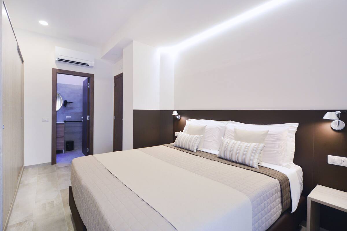 Quarto de hotel moderno com uma cama grande, uma mesinha de cabeceira e um criado-mudo.