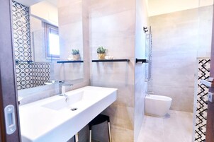 Casa de banho moderna com lavatório branco, espelho, sanita e chuveiro. Existem estantes com objetos decorativos e uma janela sobre o lavatório.