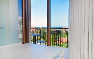 Uma varanda com mesa e cadeiras, vista para uma área costeira com edifícios e o mar.