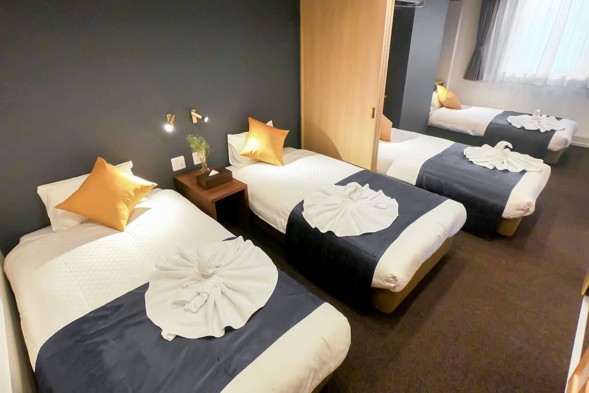Quarto de hotel com três camas, cada uma com colcha branca e azul escura, toalha branca e pequena mesa de cabeceira com abajur e um vaso.