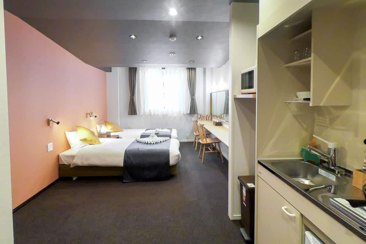 Quarto de hotel com cama, mesas de cabeceira, escrivaninha, cadeira, televisão e cozinha compacta.