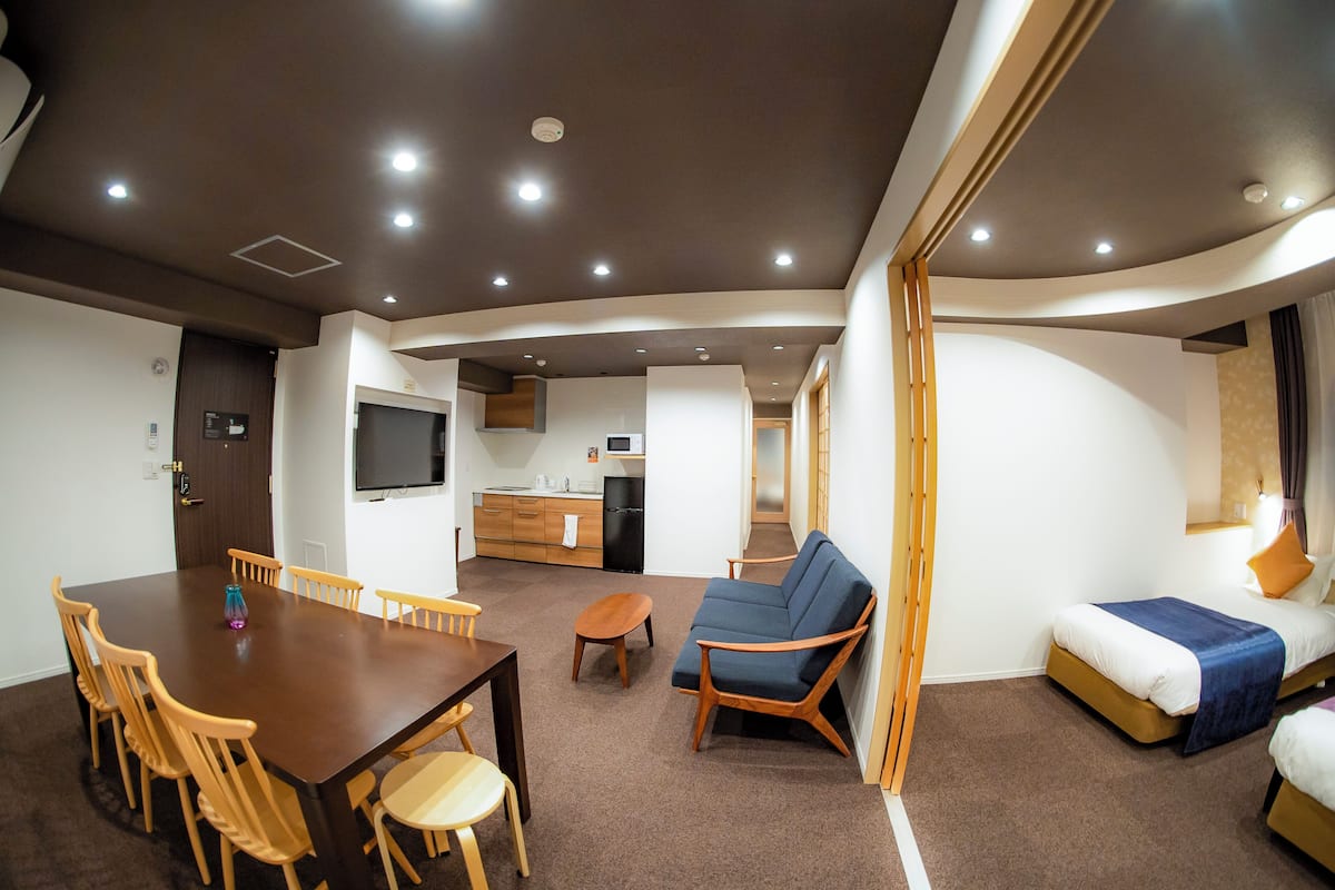 Quarto de hotel moderno com área de refeições, sofá, cama e cozinha compacta.