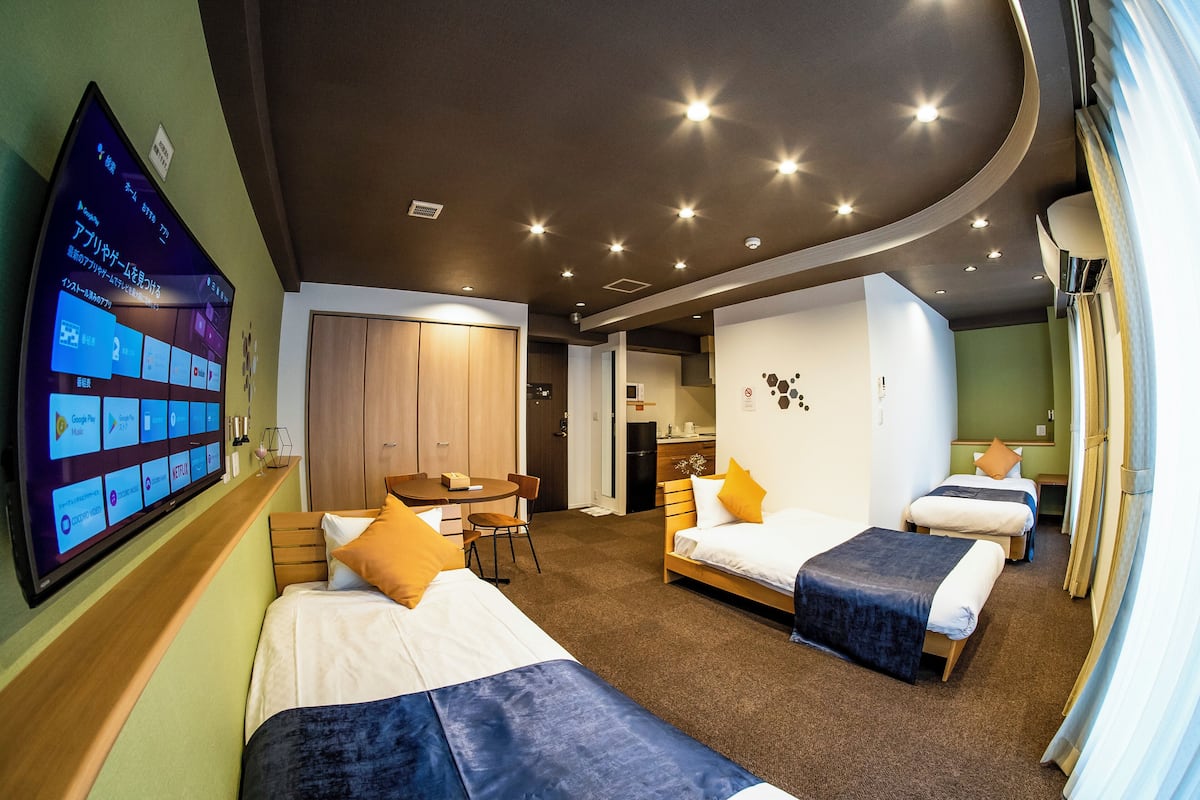 Quarto de hotel com três camas, uma televisão fixada na parede, uma pequena mesa de jantar e uma cozinha compacta.