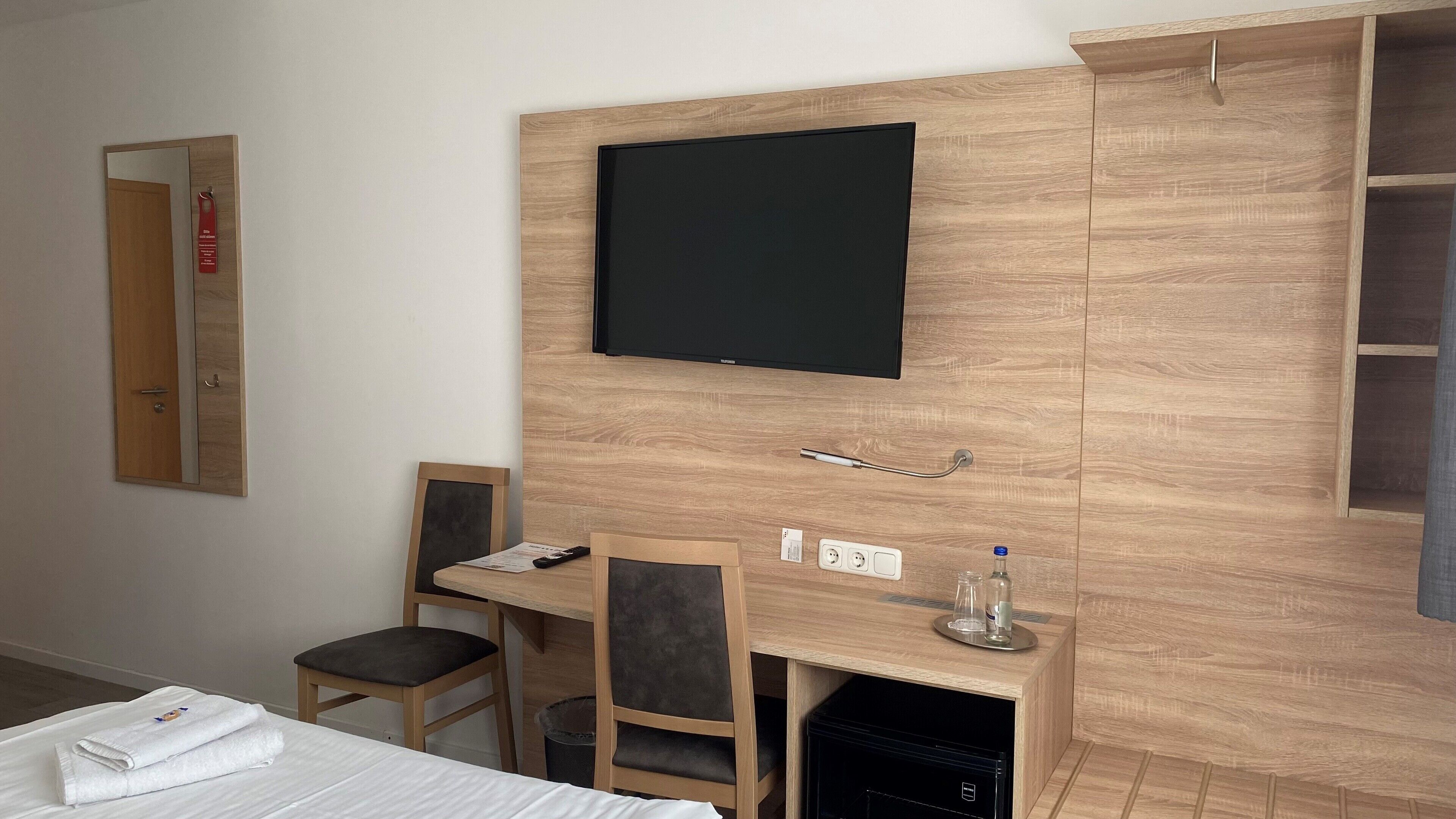Quarto de hotel com cama, escrivaninha com cadeira, televisão na parede e espelho.