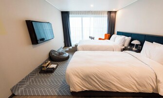 Quarto de hotel com duas camas, uma televisão, uma secretária com abajur e uma janela com cortinas.