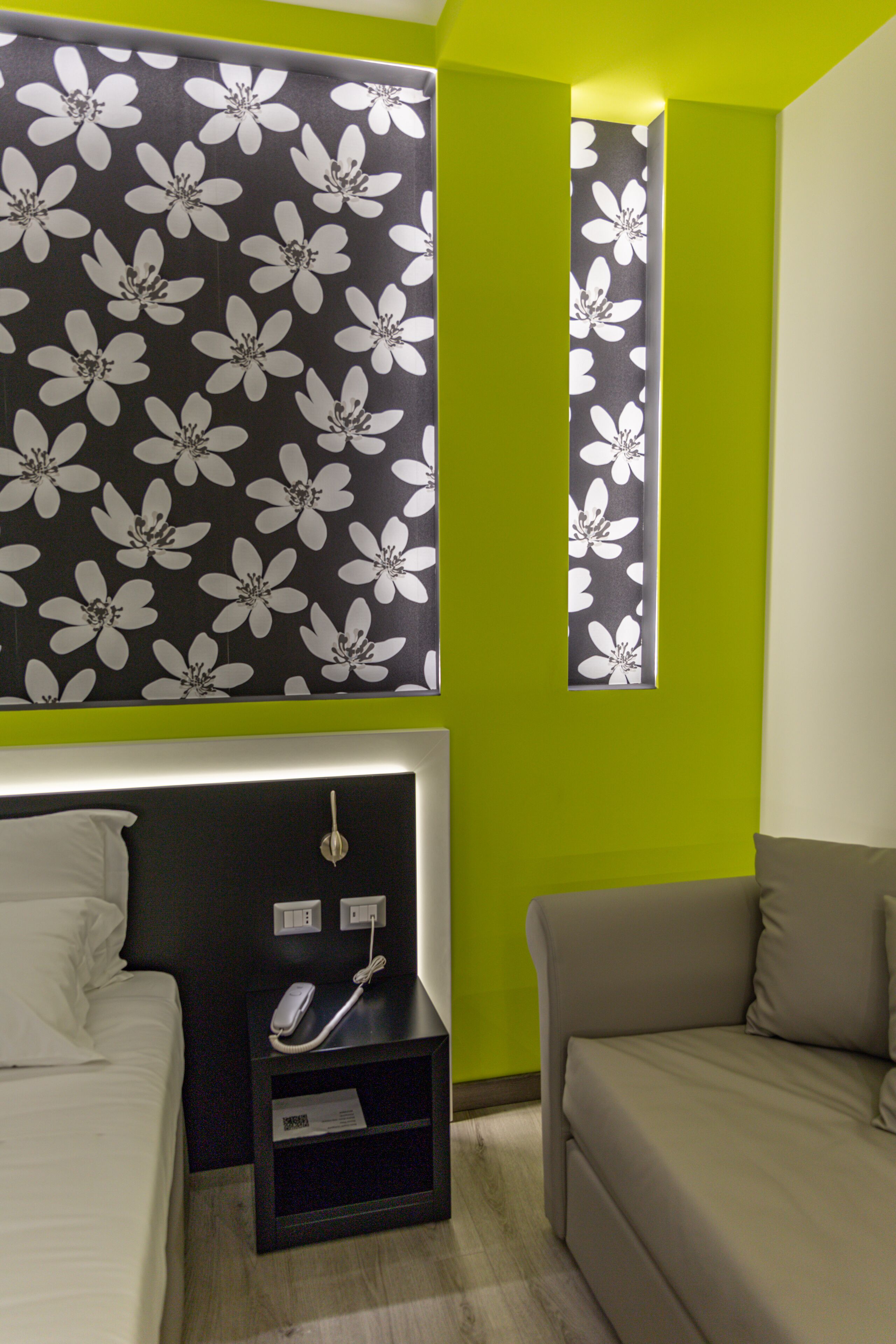 Quarto de hotel com cama, mesa de cabeceira com telefone, prateleira pequena, sofá e janela com padrão floral.